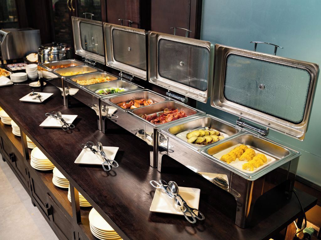 Disposição de buffet com diversas estações de comida, cada uma com uma bandeja de metal e uma tampa de vidro.