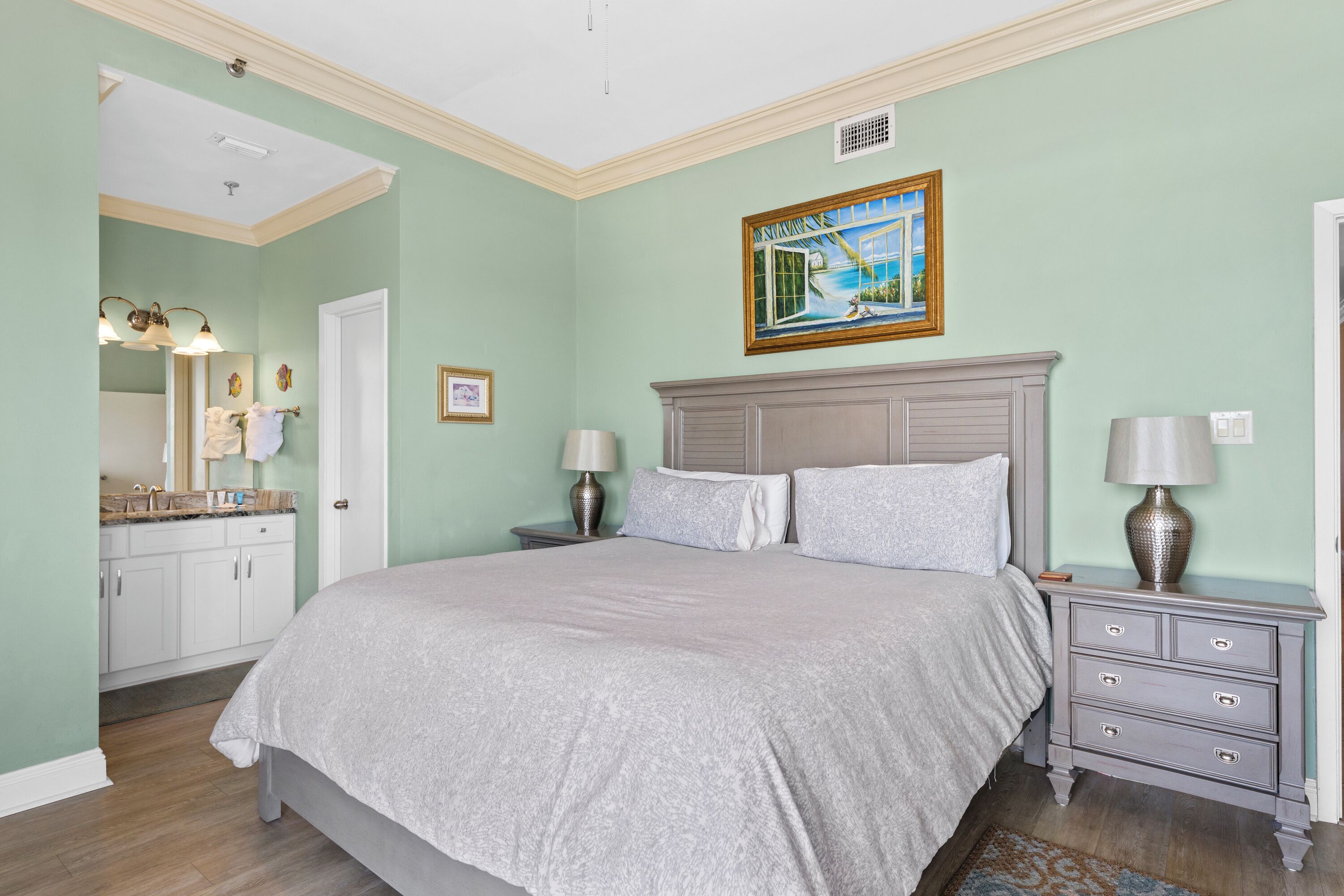 Quarto com uma cama grande, mesa de cabeceira com luminárias, cômoda e banheiro com toucador, além de um quadro na parede.