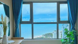 Quarto com uma grande janela com vista para a costa, uma mesa de madeira, uma planta em vaso e um vaso com penas brancas.