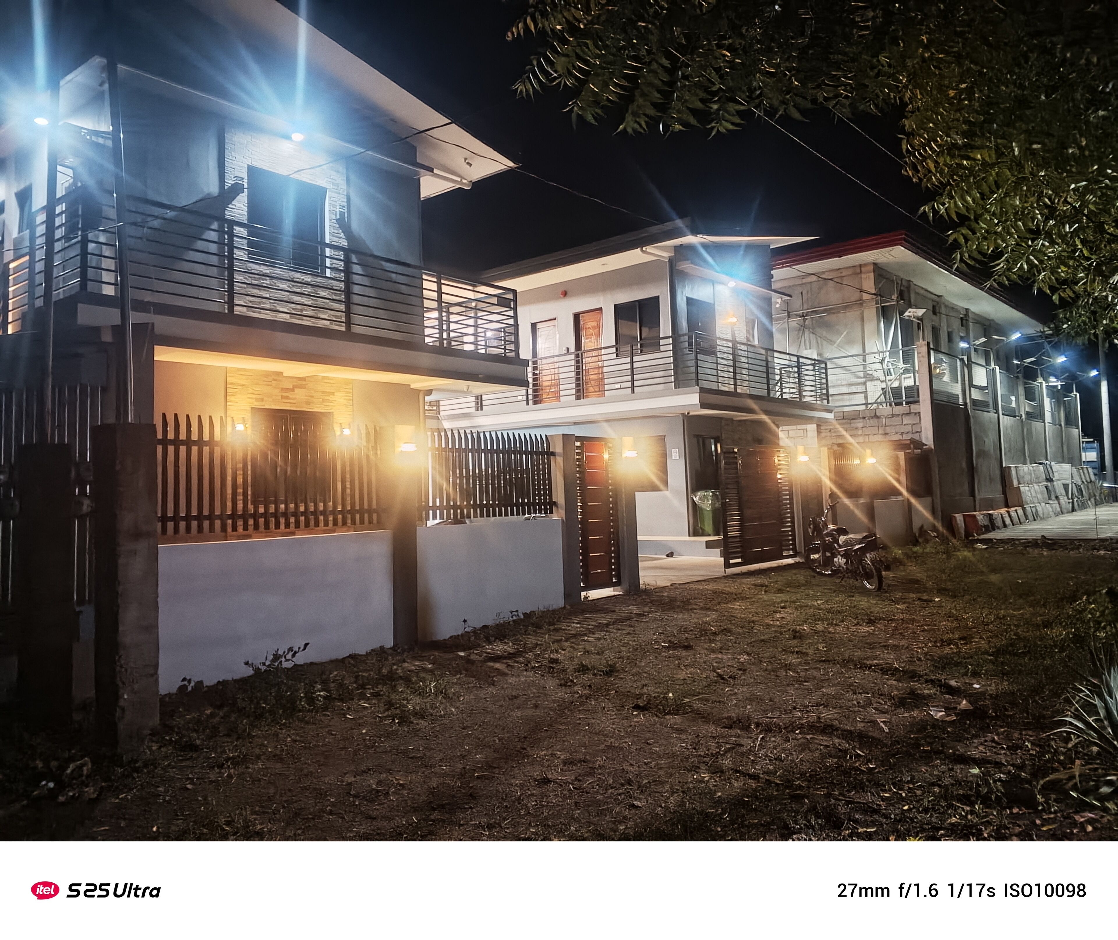 Uma casa moderna de dois andares com portão fechado, uma motocicleta estacionada na frente e uma entrada de garagem bem iluminada.