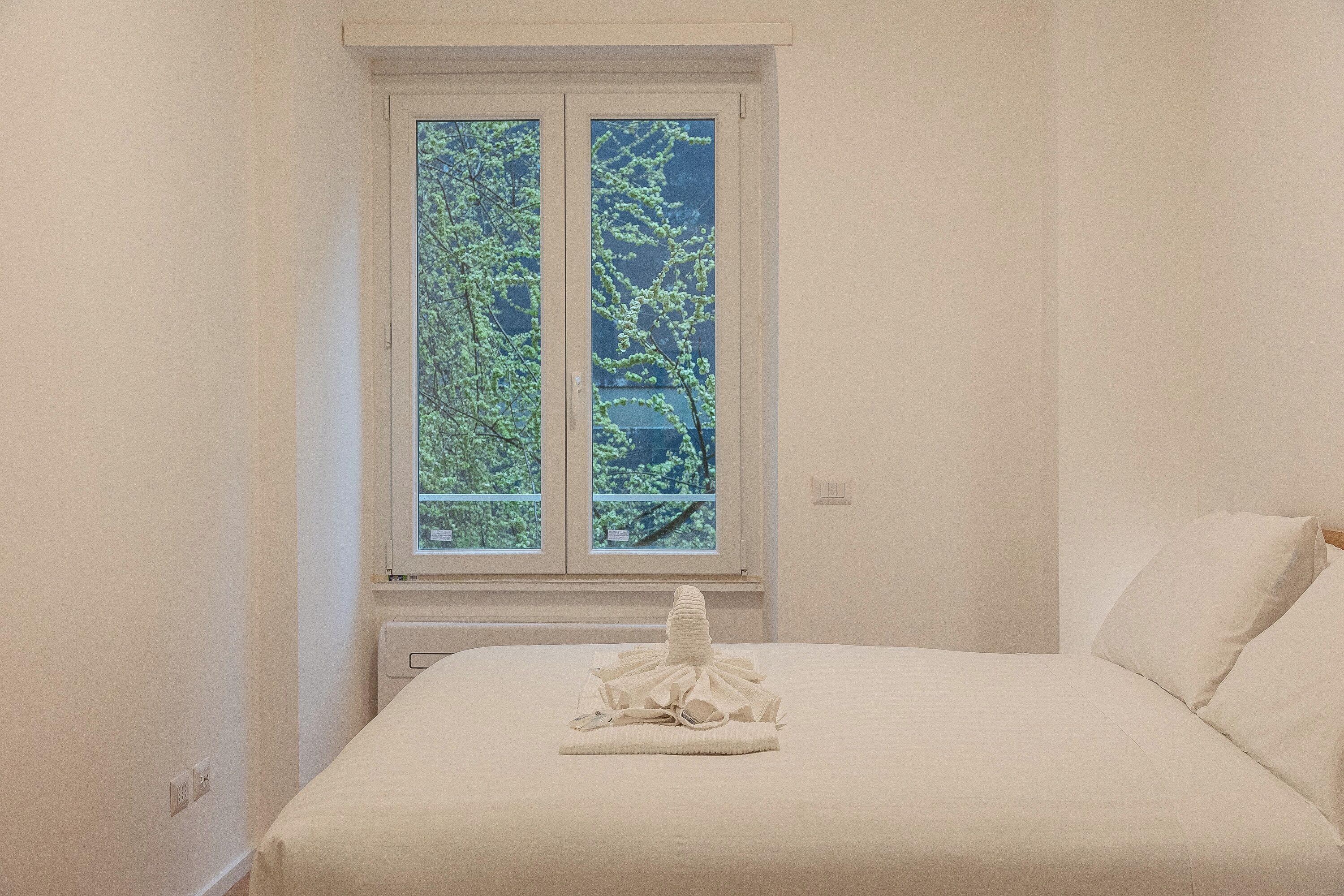 Quarto com uma janela ampla, cama com roupa de cama branca e toalha branca sobre a cama.