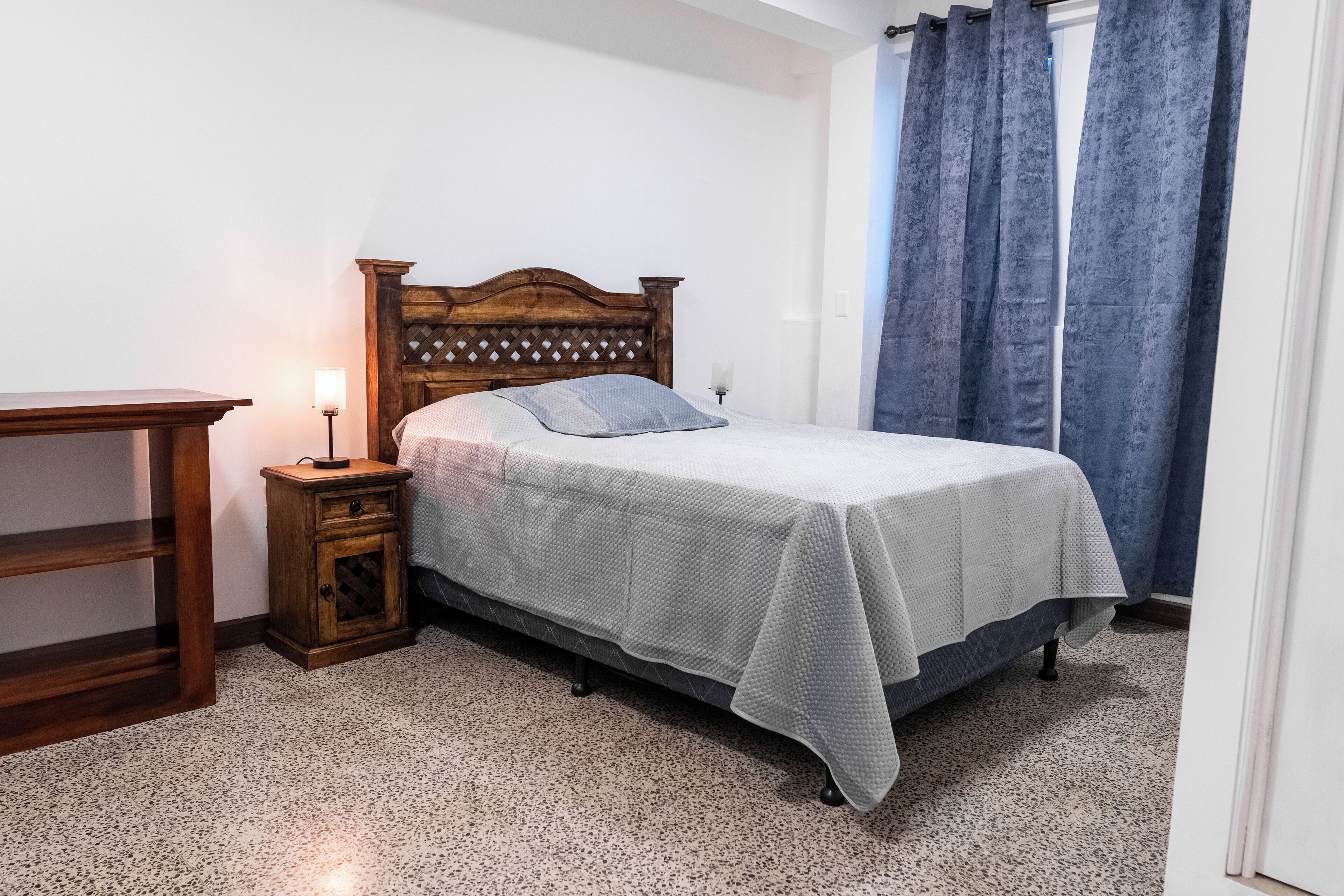Quarto com cabeceira de madeira, uma cama de solteiro, mesa de cabeceira com abajur e janela com cortinas.