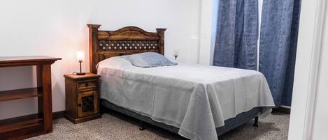 Quarto com cabeceira de madeira, uma cama de solteiro, mesa de cabeceira com abajur e janela com cortinas.