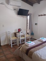 Quarto com cama, duas cadeiras, mesa, ar-condicionado, televisão, janela com cortinas e um esfregão.