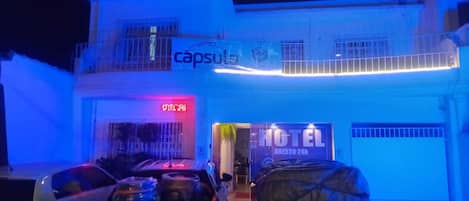 Hotel com placa indicando serviço 24 horas, um edifício de dois andares e dois carros estacionados na frente.