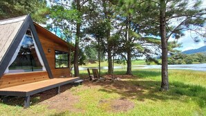 Uma cabana de madeira moderna em formato triangular, com uma janela de vidro, rodeada por árvores e vista para um lago.