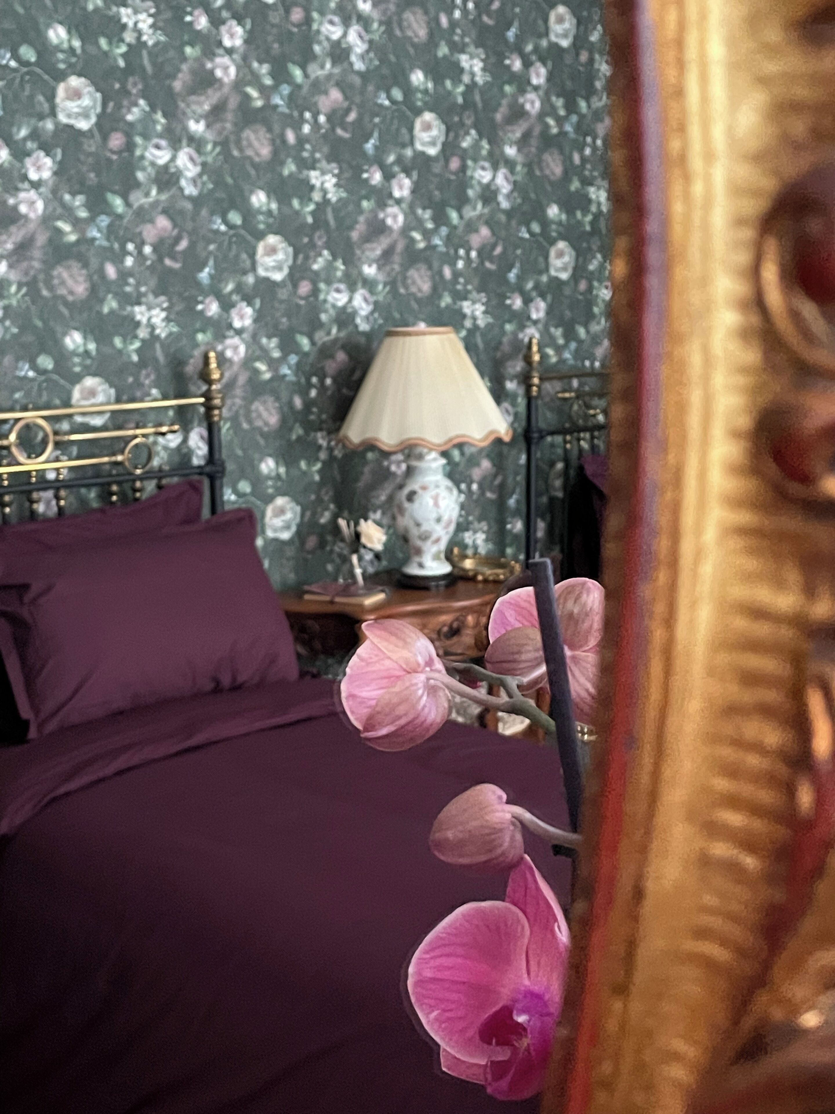 Quarto com cama, mesa de cabeceira, abajur e papel de parede floral.