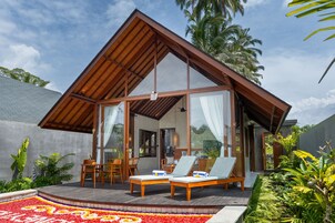 Uma villa moderna com um deck de madeira, duas poltronas de descanso, uma área de refeições e um jardim tropical.