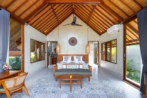 Um quarto amplo com pé direito alto, ventoinha no teto, cama com cabeceira de madeira, área de refeições com mesa e cadeiras, e grandes janelas com vista para o verde.