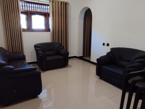 Sala de estar com três sofás pretos, uma janela com cortinas e piso em azulejos.