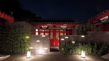 Um edifício tradicional com entrada vermelha, iluminado por luzes no chão, rodeado por bambu e arbustos.