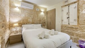 Quarto com parede de pedra, cama com roupa de cama branca, abajur, guarda-roupa de madeira e unidade de ar condicionado.