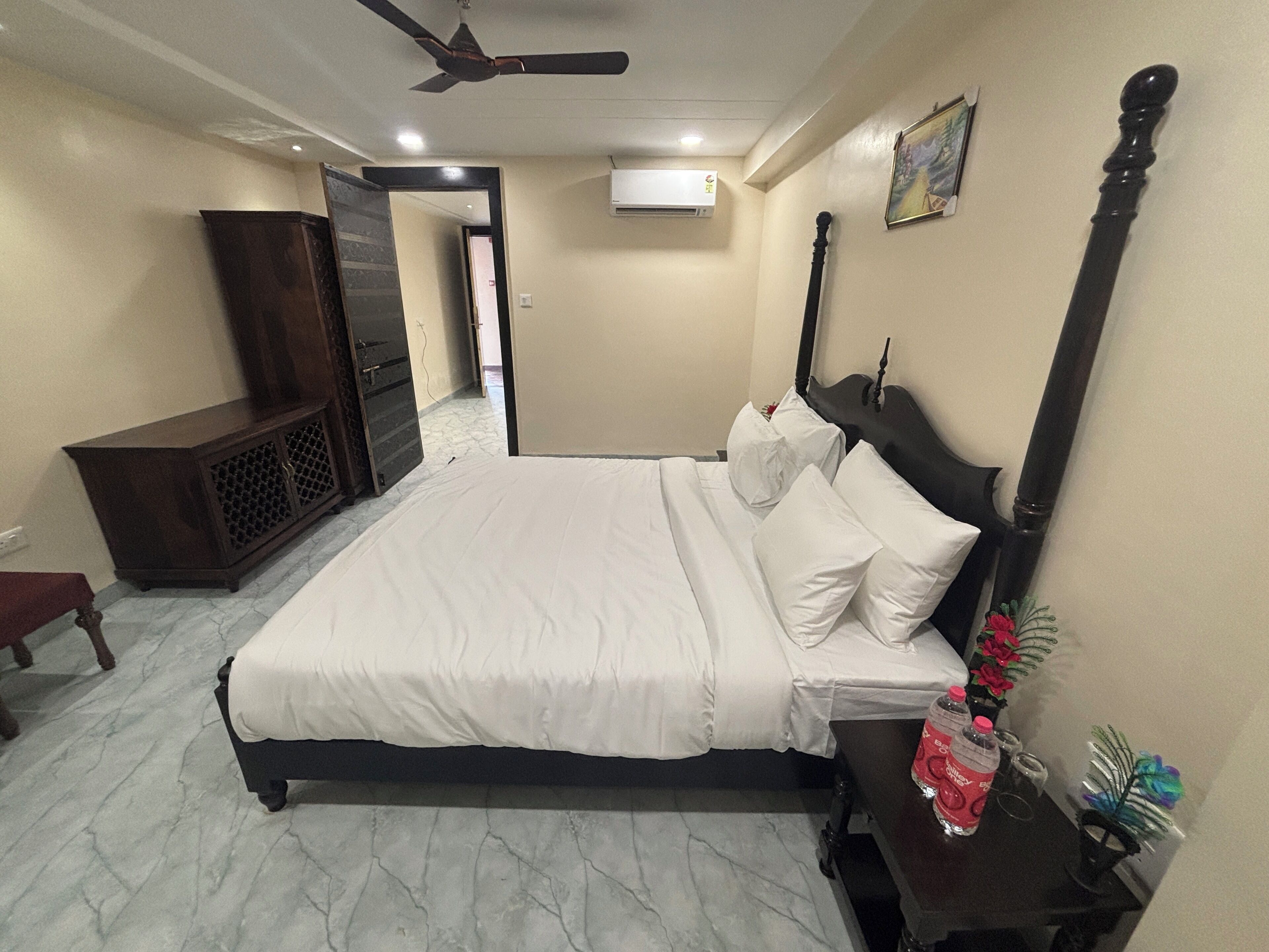 Cama bem arrumada com lençóis brancos, cabeceira de madeira escura e mesa de cabeceira com uma garrafa vermelha e copos.