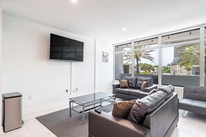 Sala de estar moderna com TV de tela plana, dois sofás cinzas, uma mesa de centro de vidro e janelas amplas com vista para palmeiras e edifícios.