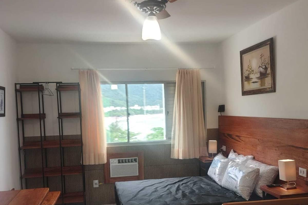 Quarto com cama, escrivaninha, cadeira, janela com cortinas, ventilador de teto e ar-condicionado fixado na parede.