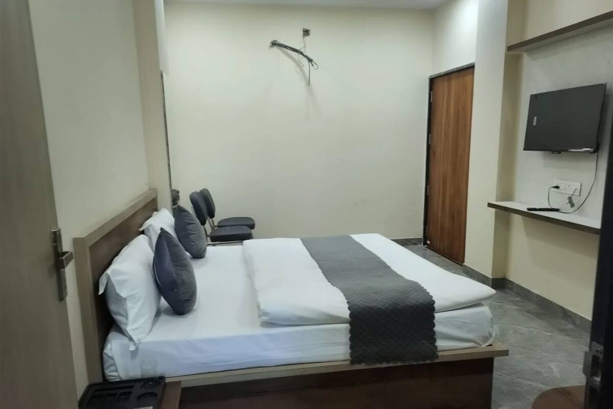 Quarto pequeno e bem arrumado com cama, almofadas, mesa de cabeceira, televisão fixada na parede e porta de madeira.
