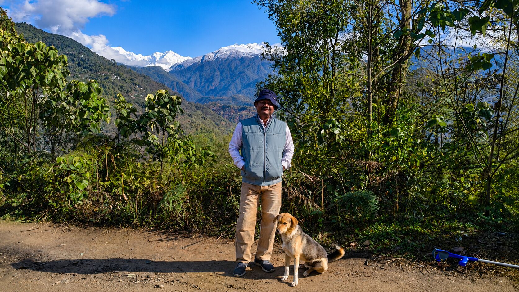 Uma pessoa está de pé com um cão numa trilha de terra, rodeada por vegetação e montanhas ao fundo.
