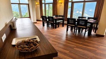 Uma área de refeições com uma mesa de madeira comprida e cadeiras, uma mesa de buffet com um prato de comida, e janelas amplas com vista para o exterior.