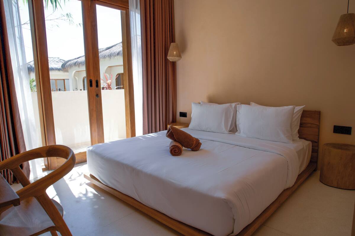 Quarto de hotel com cama, uma cadeira, mesa de cabeceira de madeira e vista através de uma porta de correr.