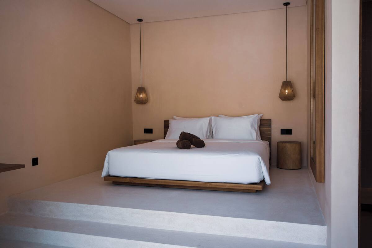 Quarto minimalista com uma cama, duas luzes pendentes e um banquinho pequeno.