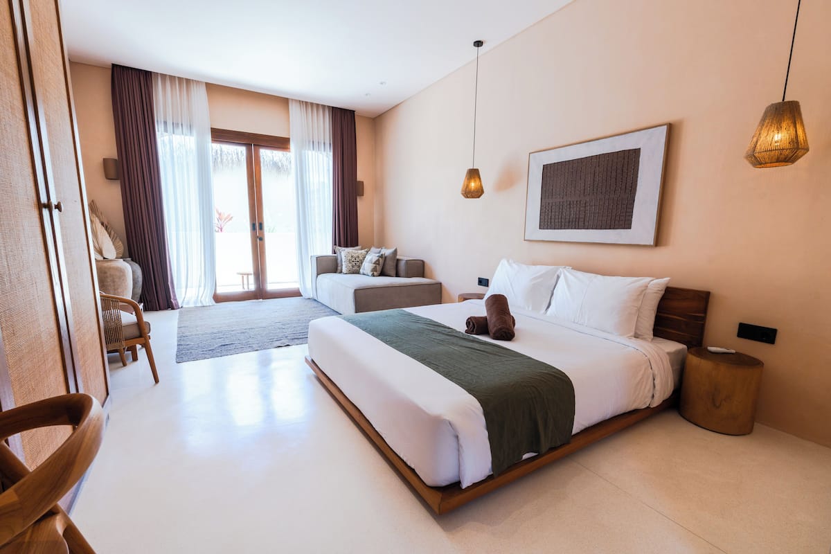 Quarto de hotel com uma cama grande, uma cama menor, um sofá, uma cadeira, uma mesinha de madeira redonda e um lustre.