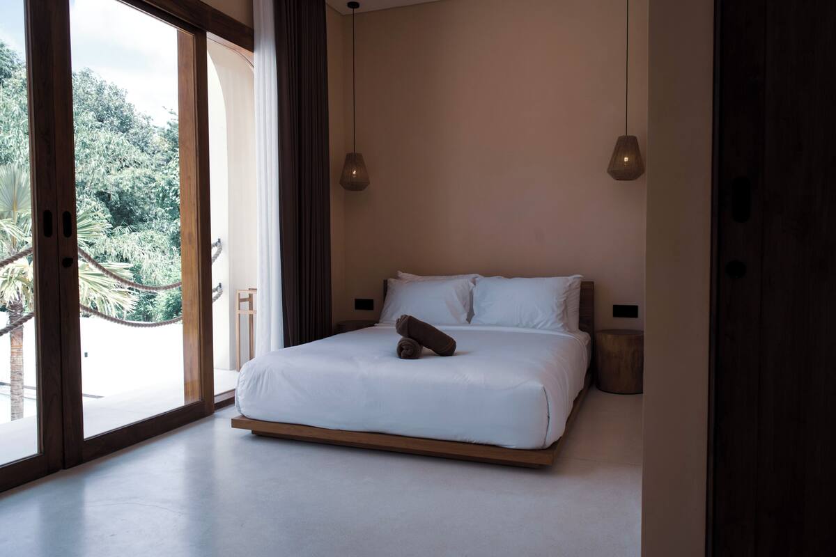 Quarto com uma cama grande, vista para o verde lá fora e duas luminárias pendentes.