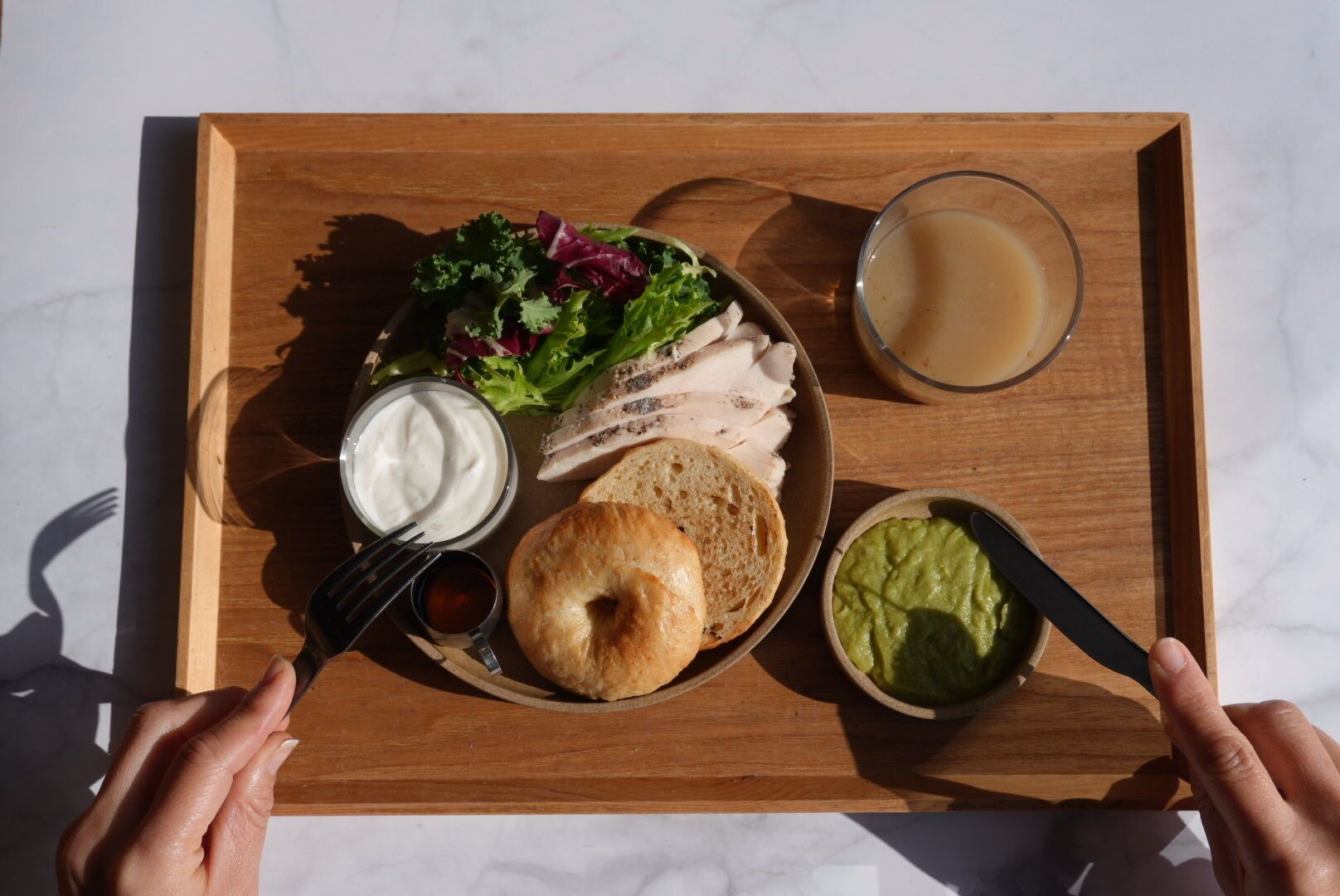Uma bandeja de madeira com uma salada, fatias de peru, um bagel e duas tigelas de molho.
