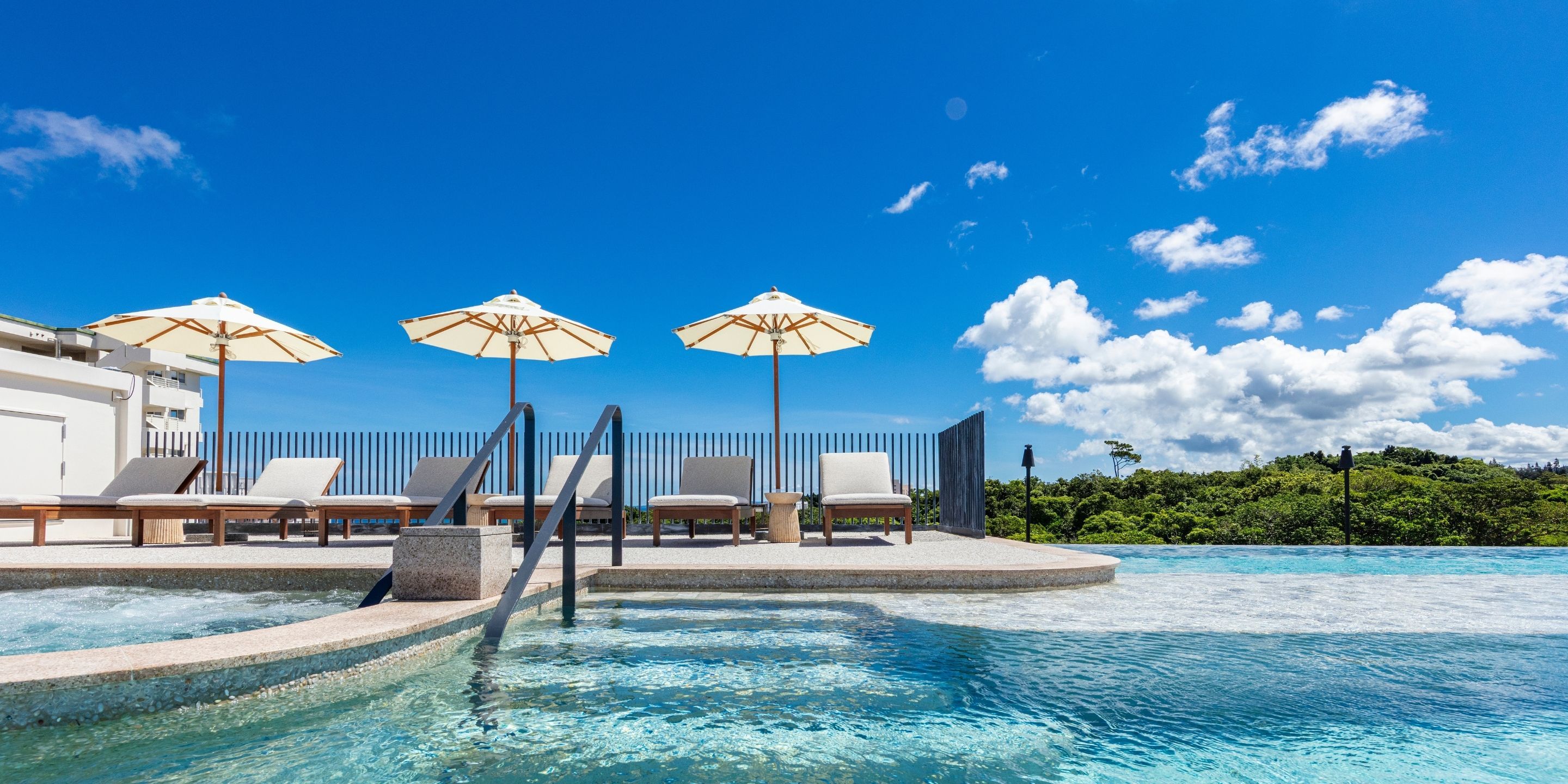 Uma área de piscina com espreguiçadeiras e guarda-sóis, cercada por uma vedação e um céu claro.