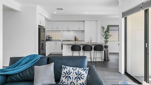 Uma sala de estar moderna com um sofá azul escuro, uma almofada decorativa estampada e um cobertor azul. Na parte de trás, há uma área de cozinha com armários brancos, bancada preta e uma pequena ilha com banquetas.