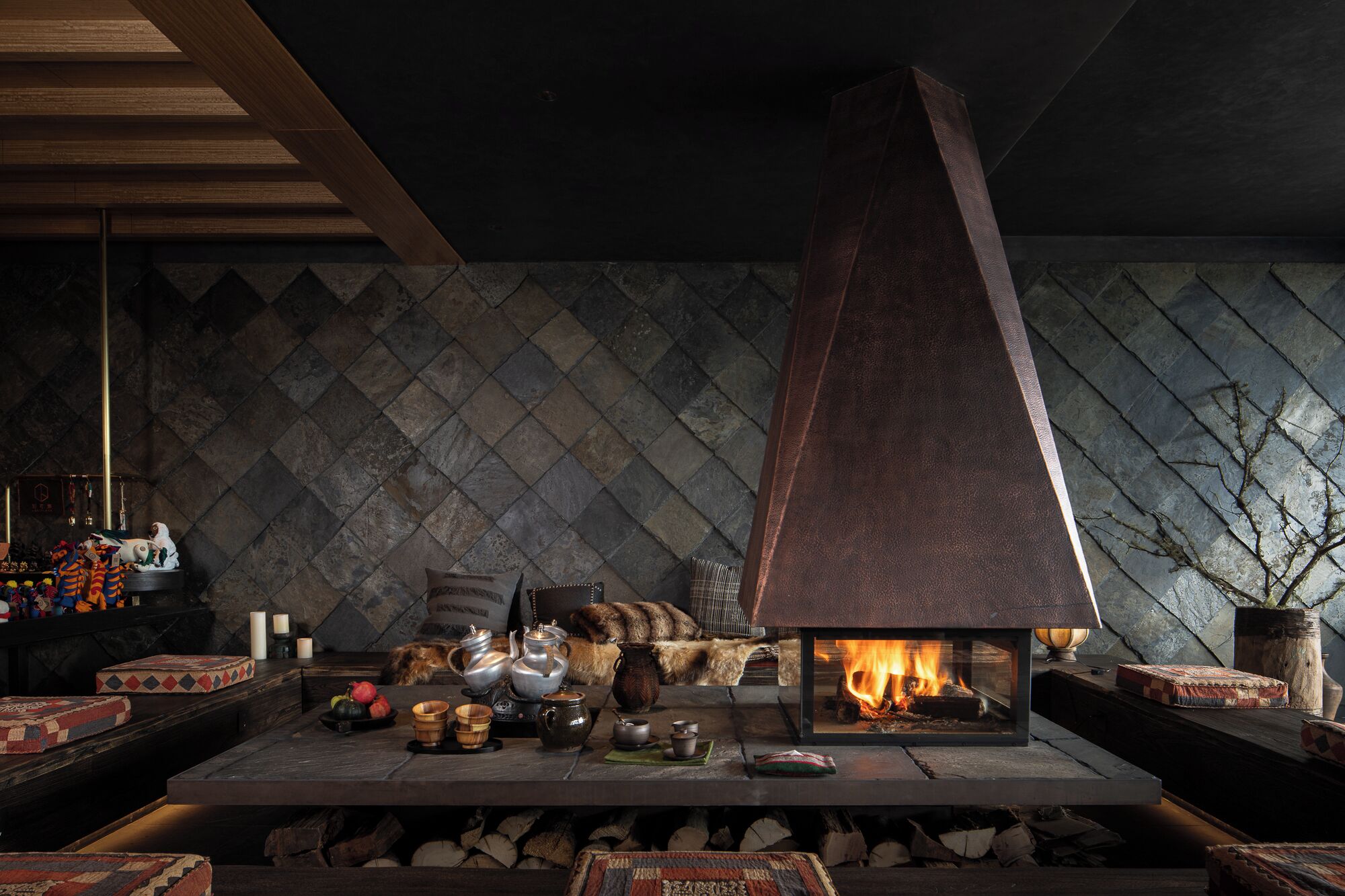 Uma área aconchegante com lareira, parede de pedra, um banco de madeira e diversos objetos decorativos.