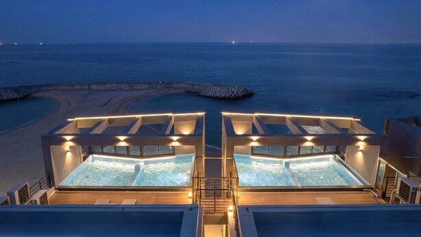 Uma piscina infinita no terraço com duas banheiras de hidromassagem, um deck de madeira e vista para o oceano ao entardecer.
