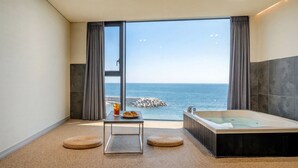 Uma casa de banho moderna com uma banheira grande, uma pequena mesa com bebida e pastelaria, e vista para o mar.