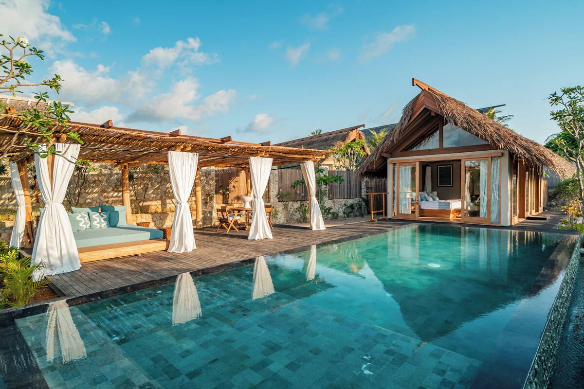 Uma área na piscina com deck de madeira, estrutura com telhado de palha e sala envidraçada.