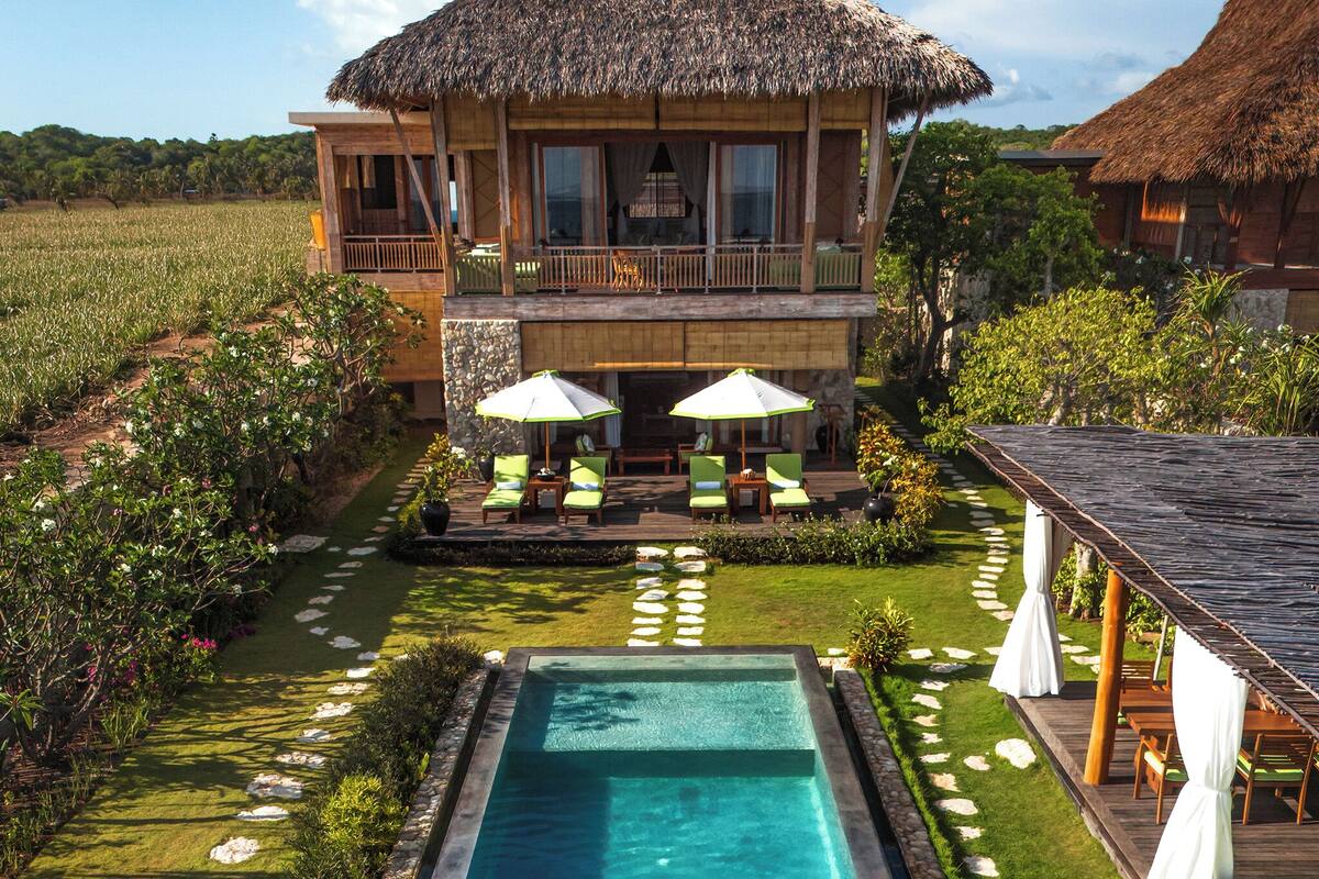 Um edifício de dois andares com telhado de palha, piscina e área de estar externa.