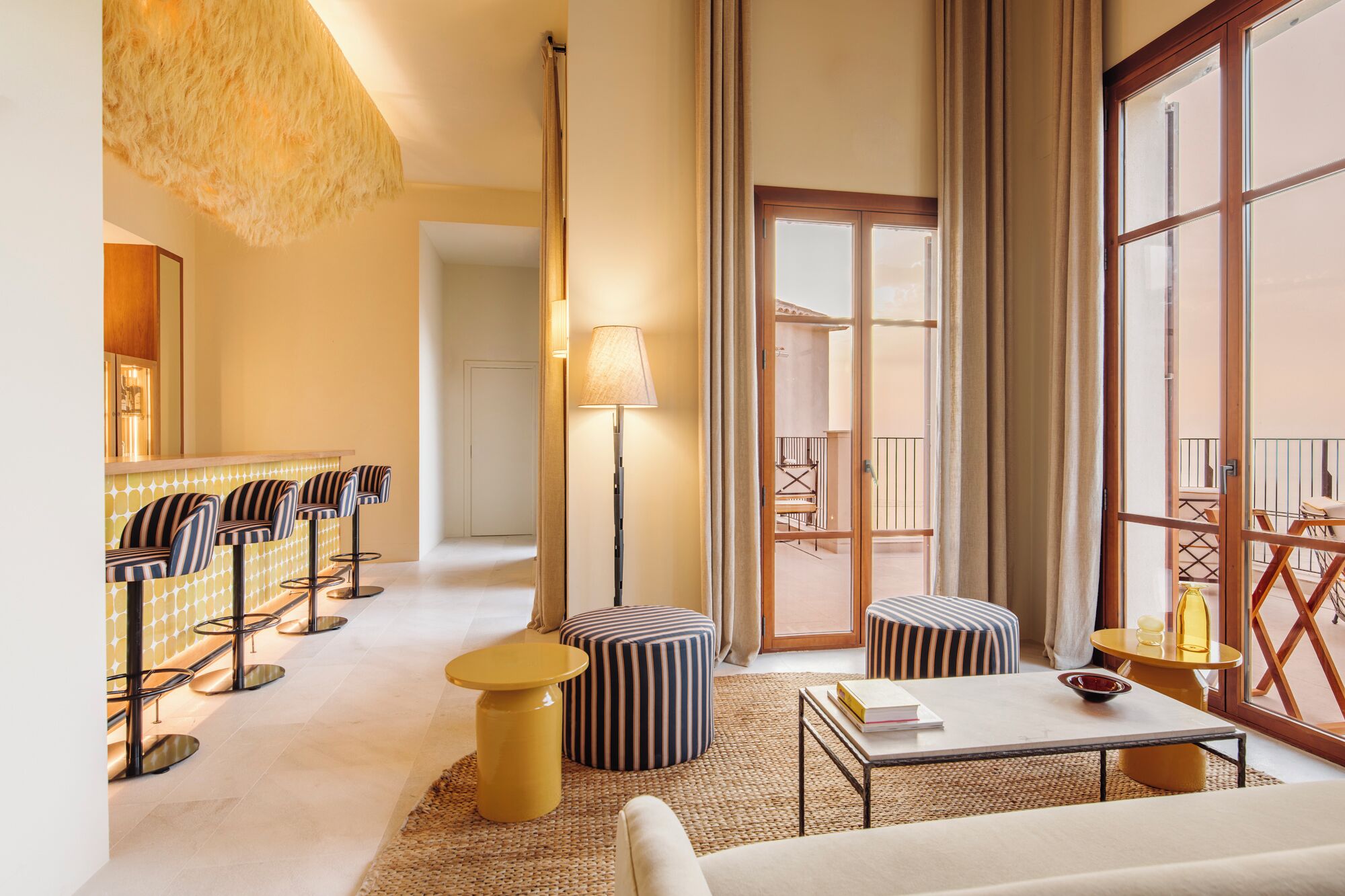 Quarto de hotel com varanda, poltronas listradas, uma mesa redonda amarela e um sofá.