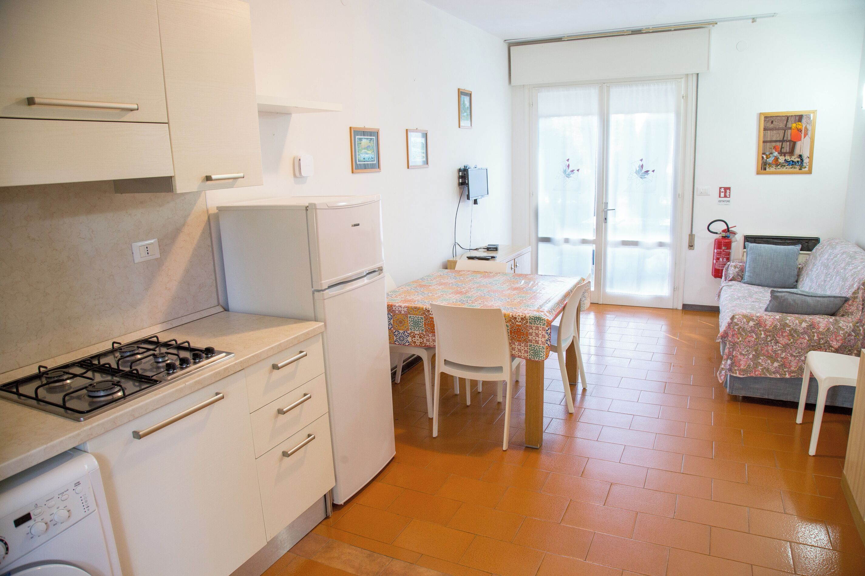 Uma cozinha compacta com fogão a gás, geladeira e máquina de lavar. Há uma mesa de jantar com cadeiras e um sofá na sala de estar.