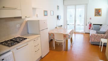 Uma cozinha compacta com fogão a gás, geladeira e máquina de lavar. Há uma mesa de jantar com cadeiras e um sofá na sala de estar.