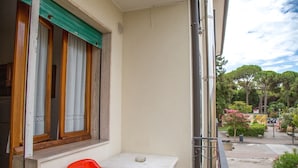 Uma varanda com uma mesa branca e uma cadeira vermelha, com vista para uma rua com árvores e uma faixa de pedestres.