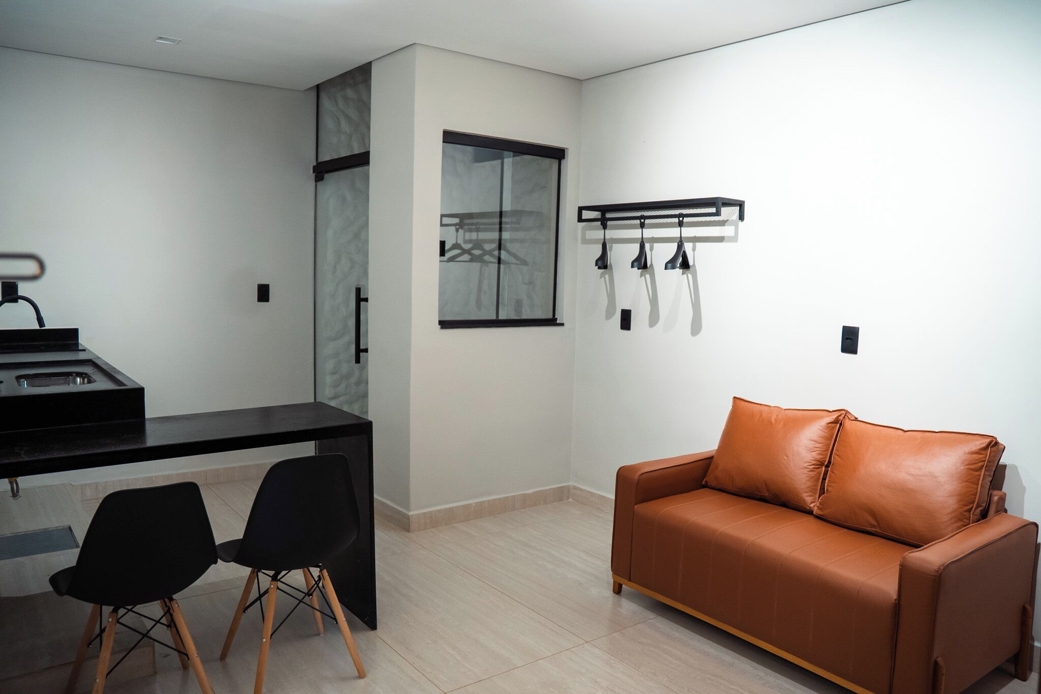 Uma sala de estar moderna com um sofá de couro marrom, uma mesa de jantar preta com duas cadeiras, uma área de cozinha com uma pia e uma janela com luminárias pendentes.