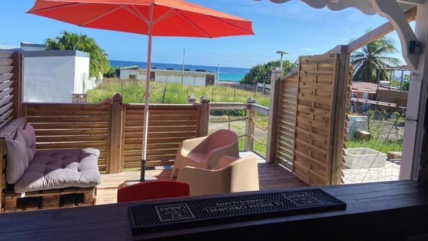 Uma área de estar coberta ao ar livre, com um guarda-sol vermelho, deck de madeira e vista para o oceano.