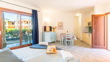 Quarto de hotel com cama, pequena área de refeições, cozinha compacta e porta de vidro deslizante que dá para área externa com plantas e um edifício ao fundo.