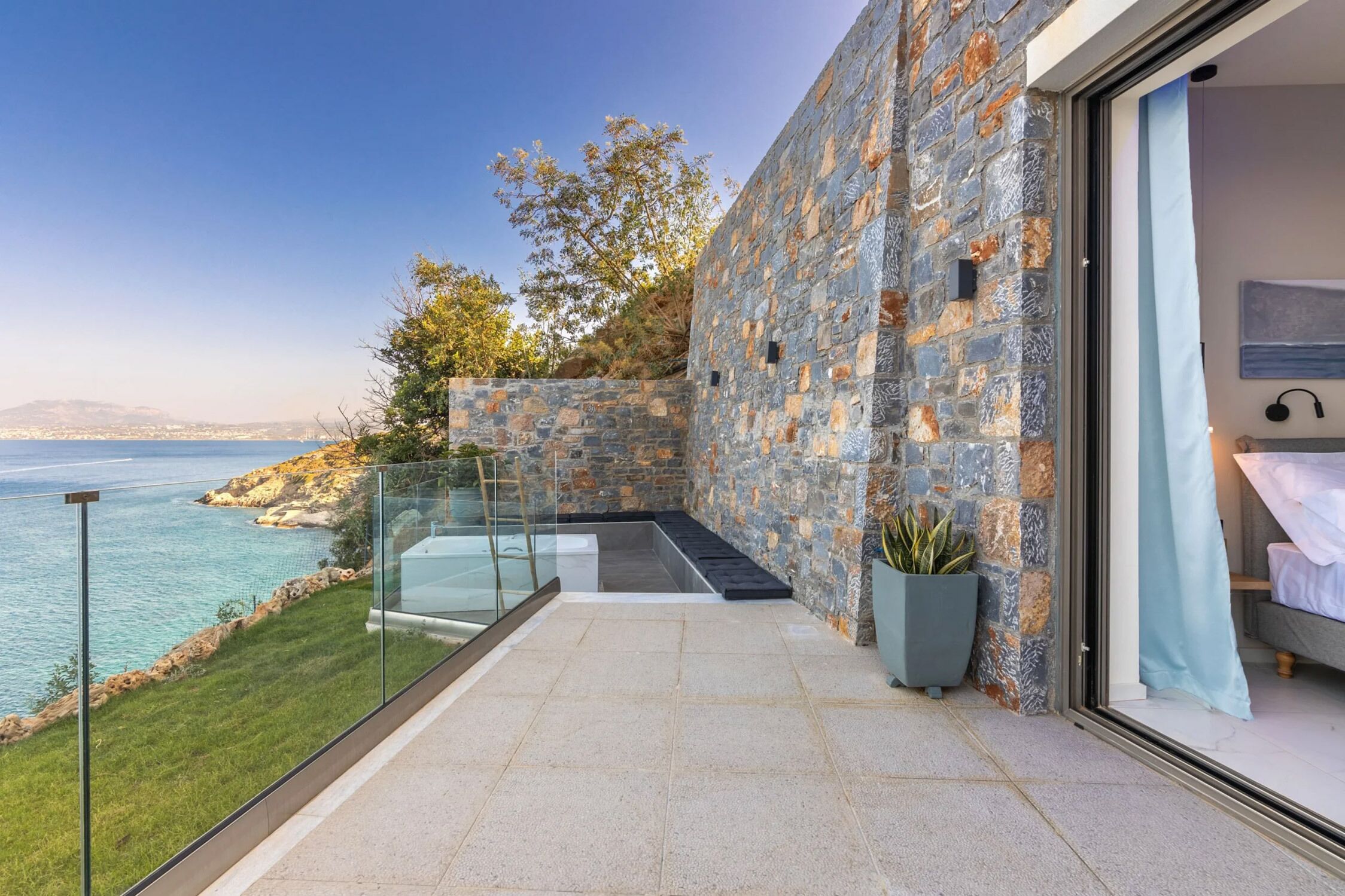 Uma varanda com parede de pedra, guarda-corpo de vidro e vista para o mar.