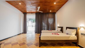Quarto de hotel com teto de madeira, duas camas com lençóis brancos, um ventilador de teto e vista para a praia através de uma grande janela.