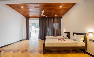 Quarto de hotel com teto de madeira, duas camas com lençóis brancos, um ventilador de teto e vista para a praia através de uma grande janela.