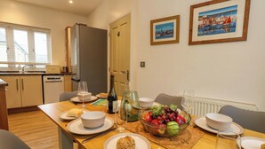 Uma área de refeições com uma mesa posta para dois, com pratos, copos, uma garrafa de vinho e uma tigela de frutas.
