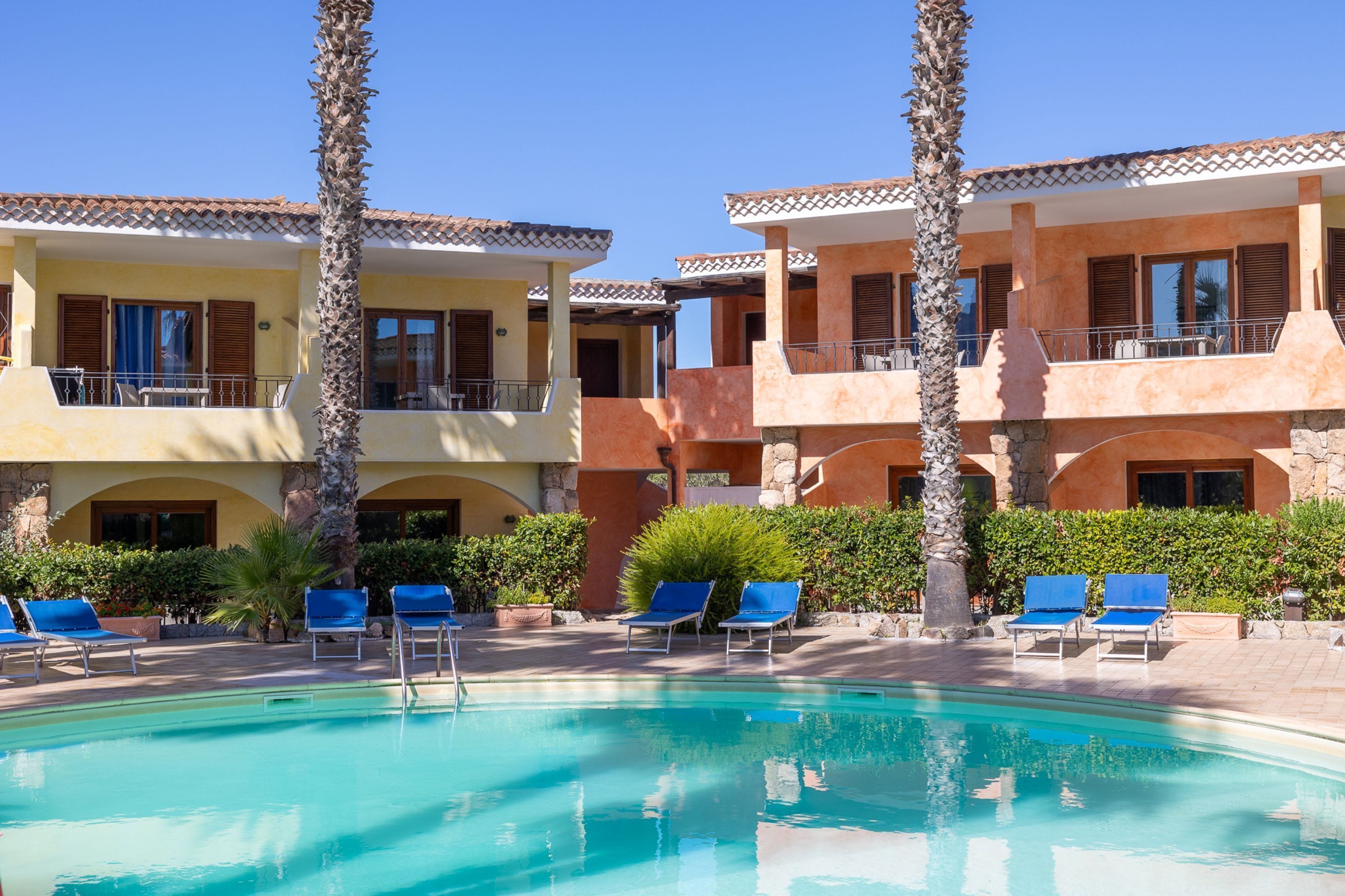 Uma área de piscina com espreguiçadeiras e piscina azul cristalina, cercada por edifícios com vários andares, varandas e janelas.