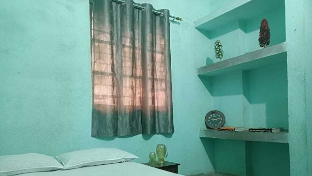 Um quarto com uma cama, mesa de cabeceira, prateleira com livros e um relógio, e uma janela com cortinas.