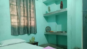 Quarto com cama, mesa de cabeceira, prateleira com objetos decorativos e janela com cortinas.