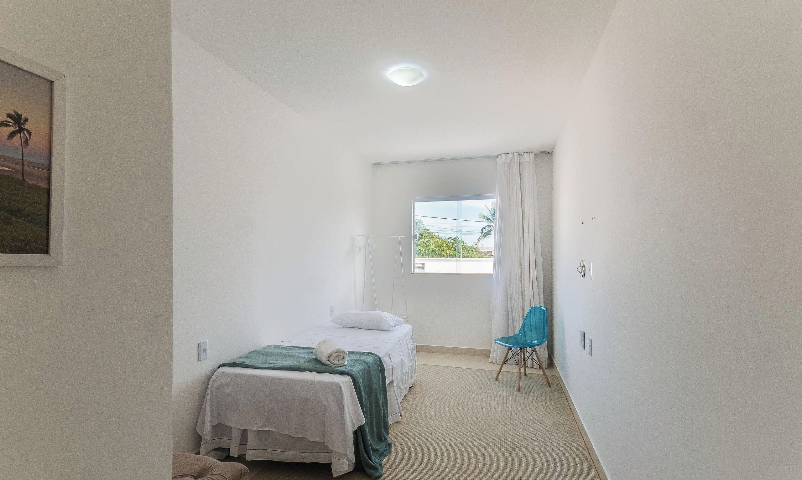 Quarto de hotel pequeno e estreito, com uma cama, uma cadeira e uma janela.