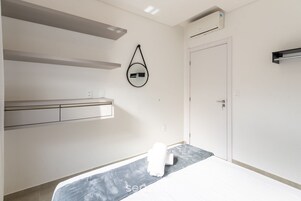 Quarto minimalista com cama, porta-toalhas fixado na parede, porta e prateleira embutida.